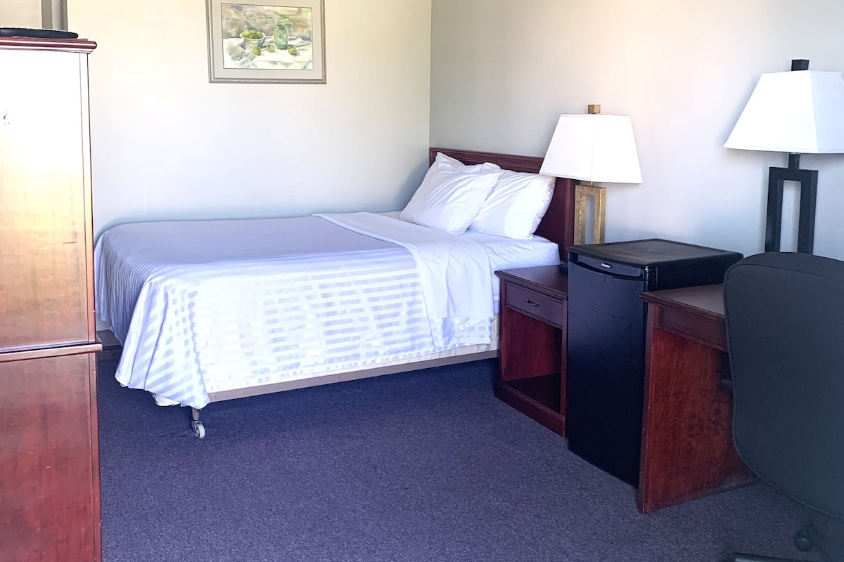 Quarto de hotel com uma cama de solteiro, duas mesas de cabeceira com luminárias, uma pequena escrivaninha e uma cadeira.