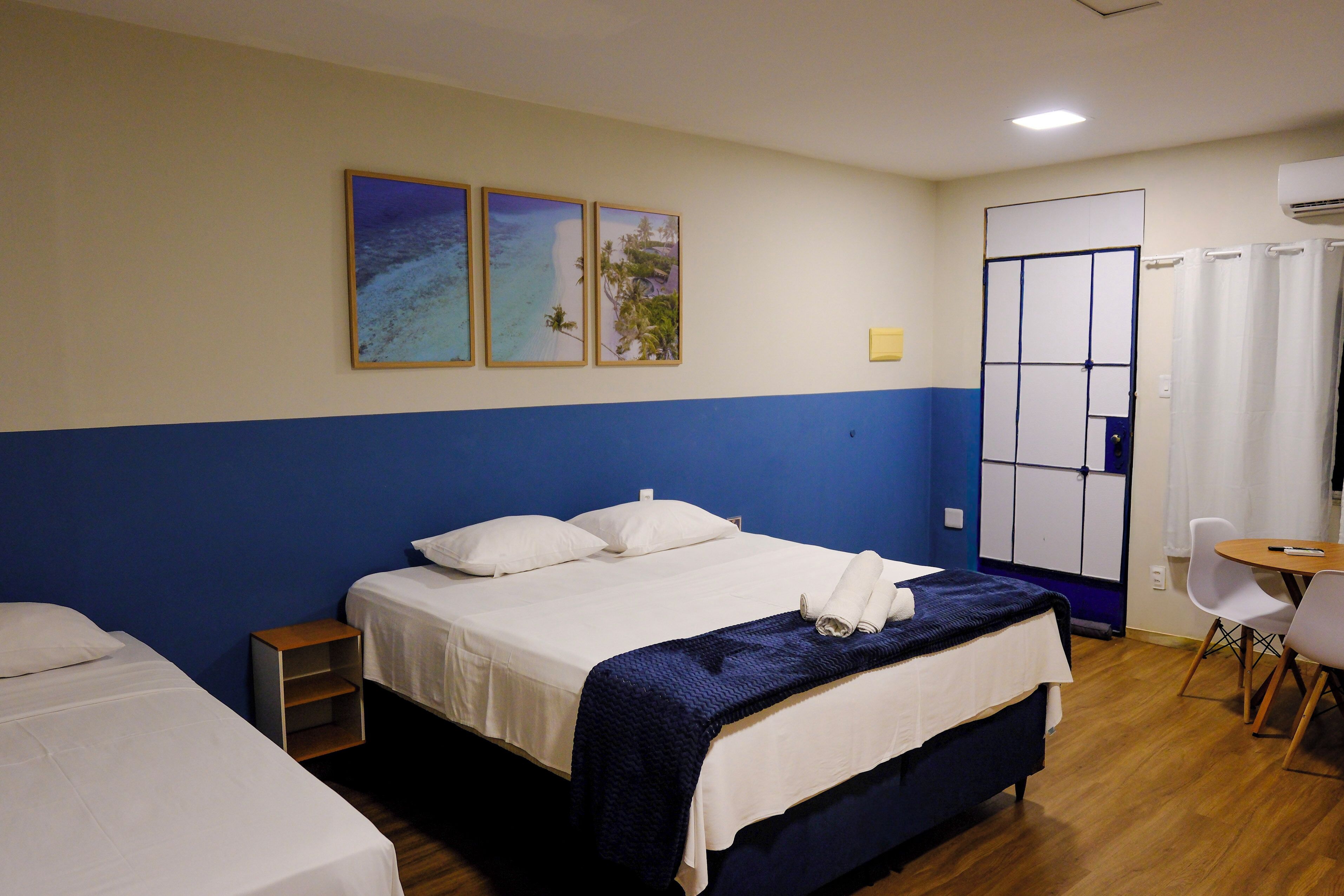 Quarto de hotel com cama, escrivaninha e cadeiras. Há três quadros na parede.