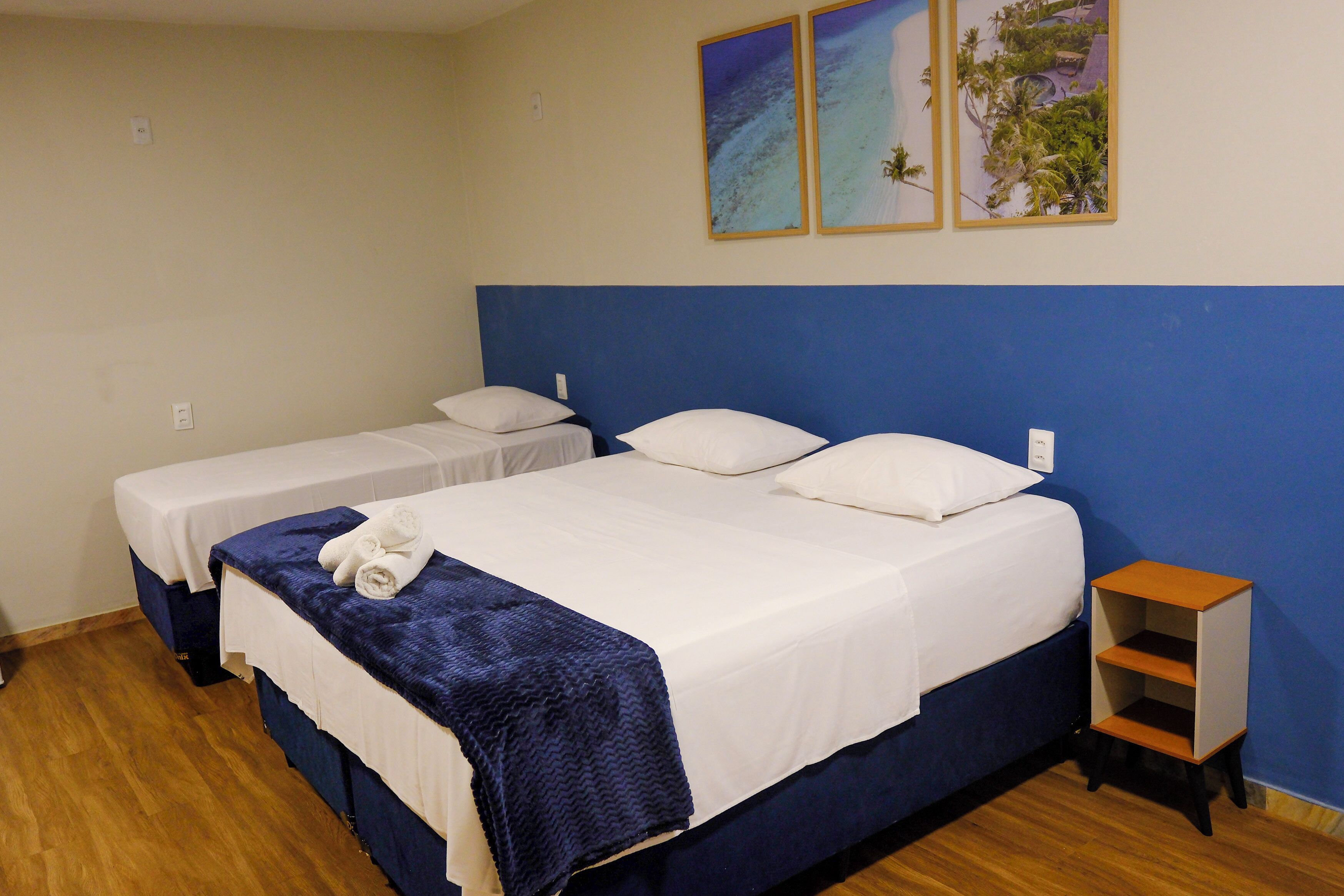 Quarto de hotel com duas camas de solteiro, cabeceira azul, roupa de cama branca e uma pequena mesa de cabeceira de madeira.