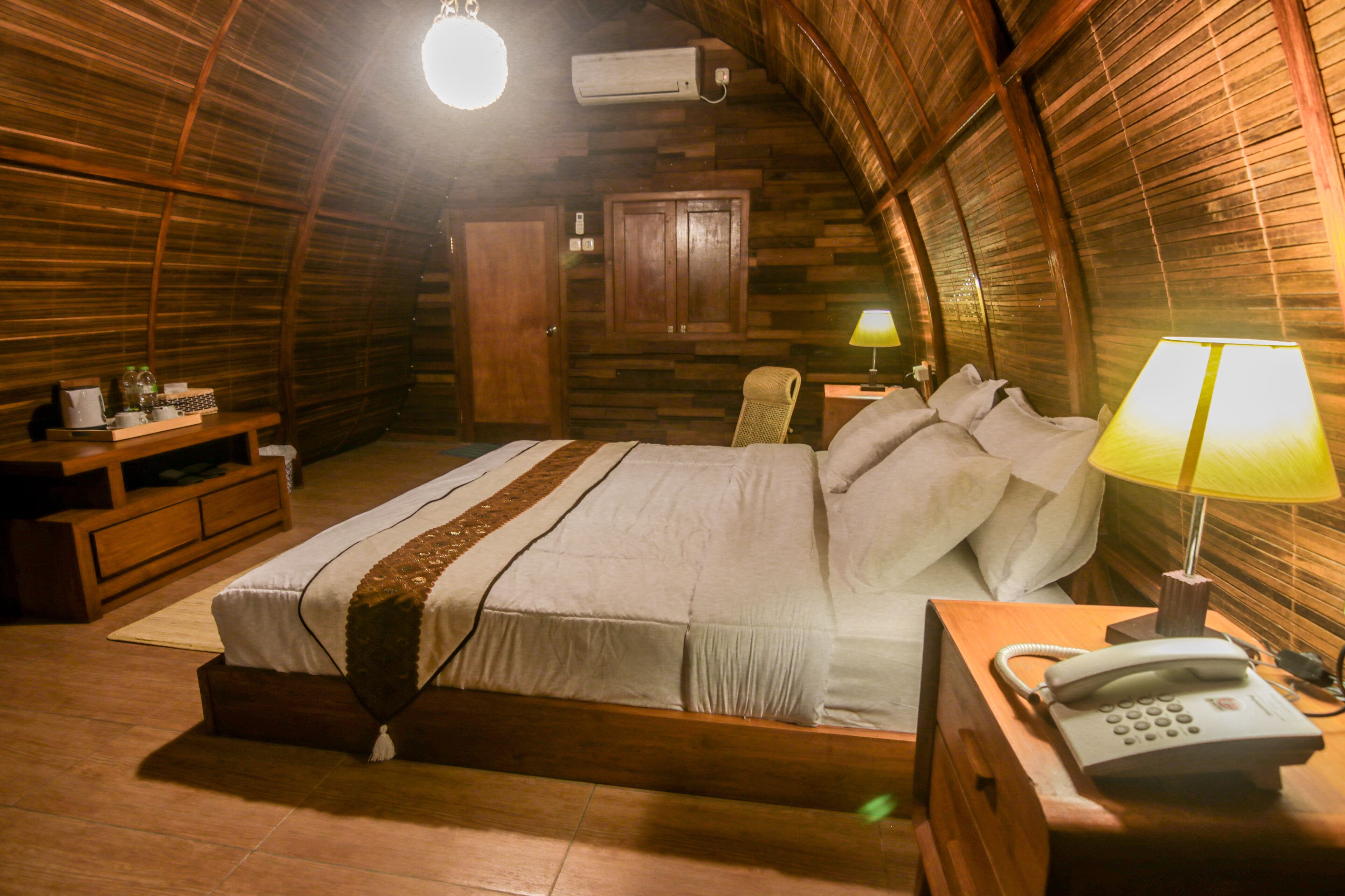 Um quarto aconchegante com piso de madeira, uma cama grande, um abajur e um telefone sobre uma mesinha de cabeceira de madeira.