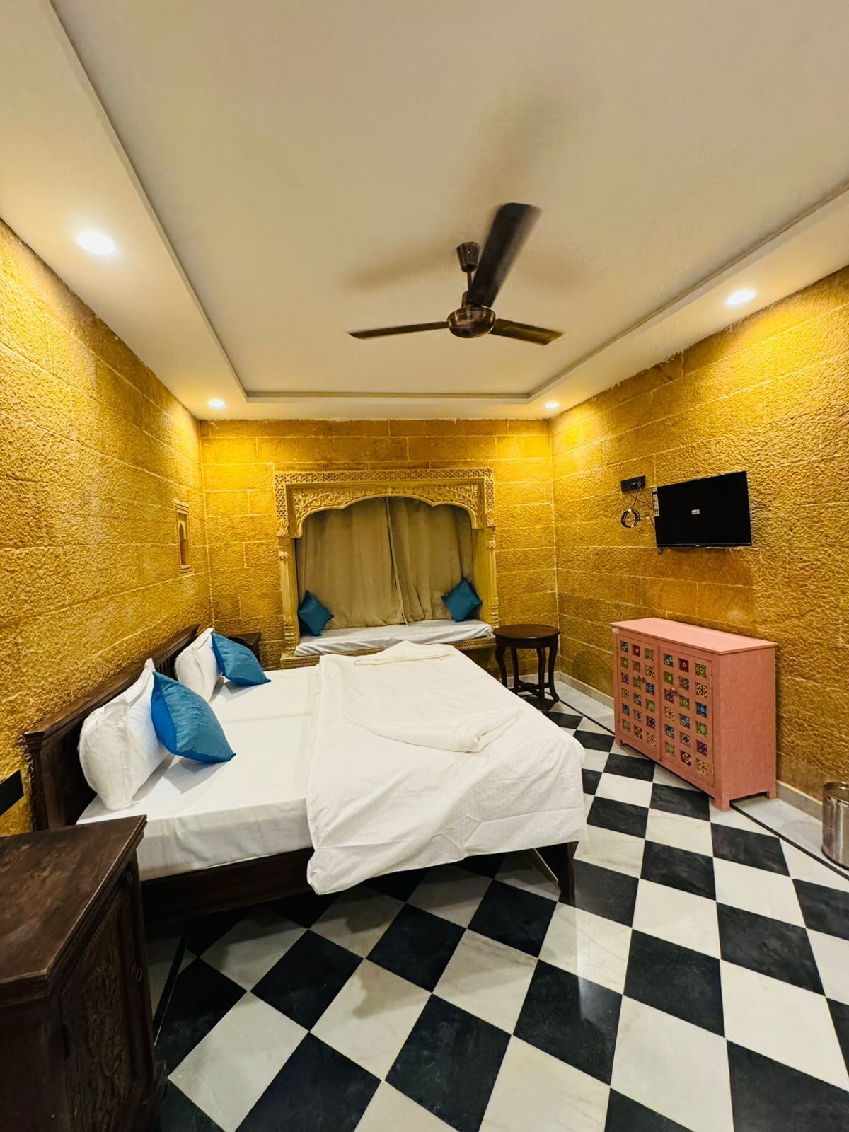 Quarto de hotel com piso xadrez, cama com lençóis brancos e almofadas azuis, ventilador de teto, TV fixada na parede e um pequeno armário rosa.