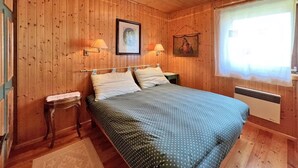 Quarto com painéis de madeira, uma cama, duas luminárias fixadas na parede, um quadro emoldurado e uma janela com cortina.