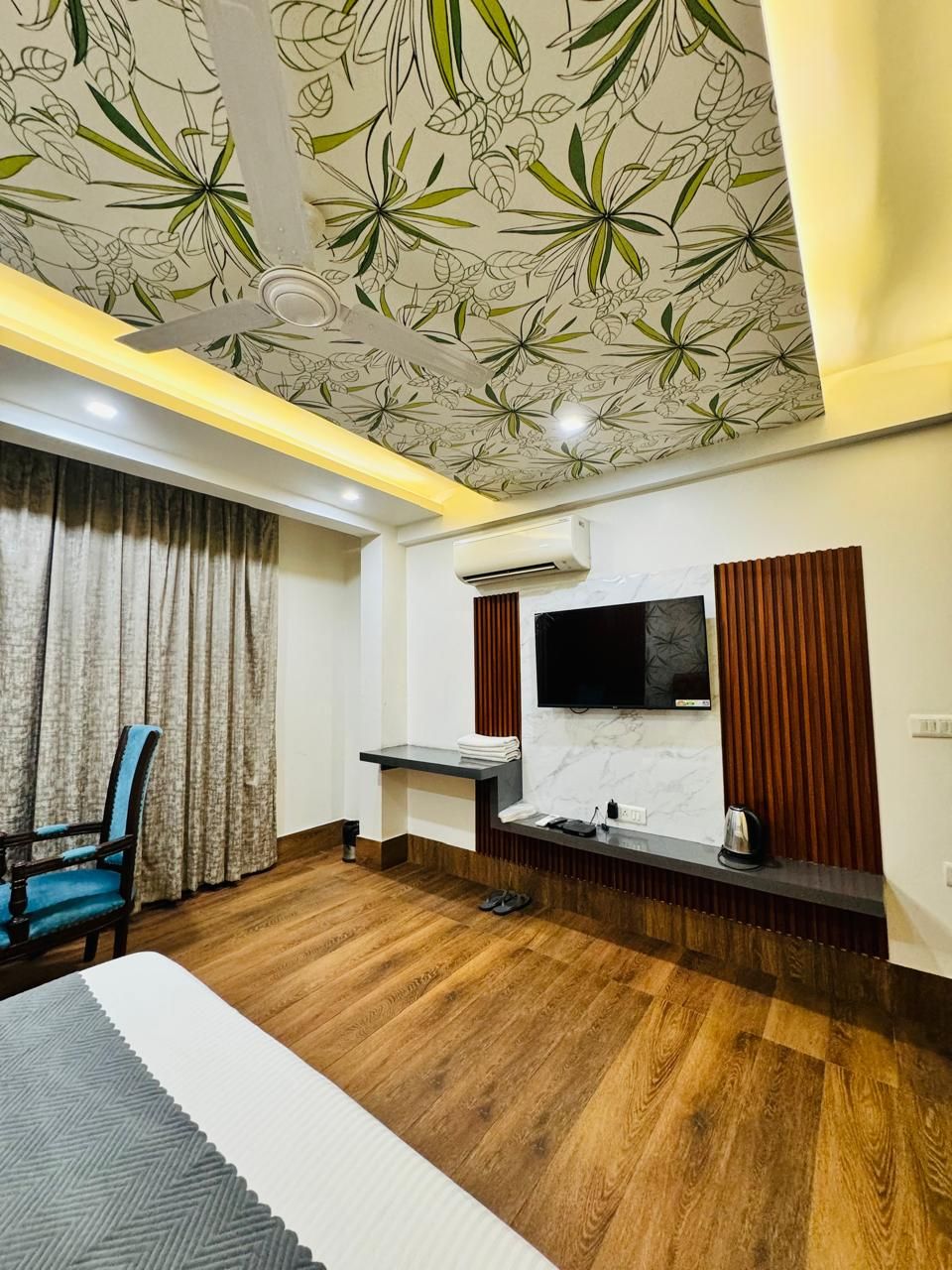 Quarto de hotel moderno com piso de madeira, uma cama, escrivaninha com televisão de tela plana e teto com padrão de folhas.