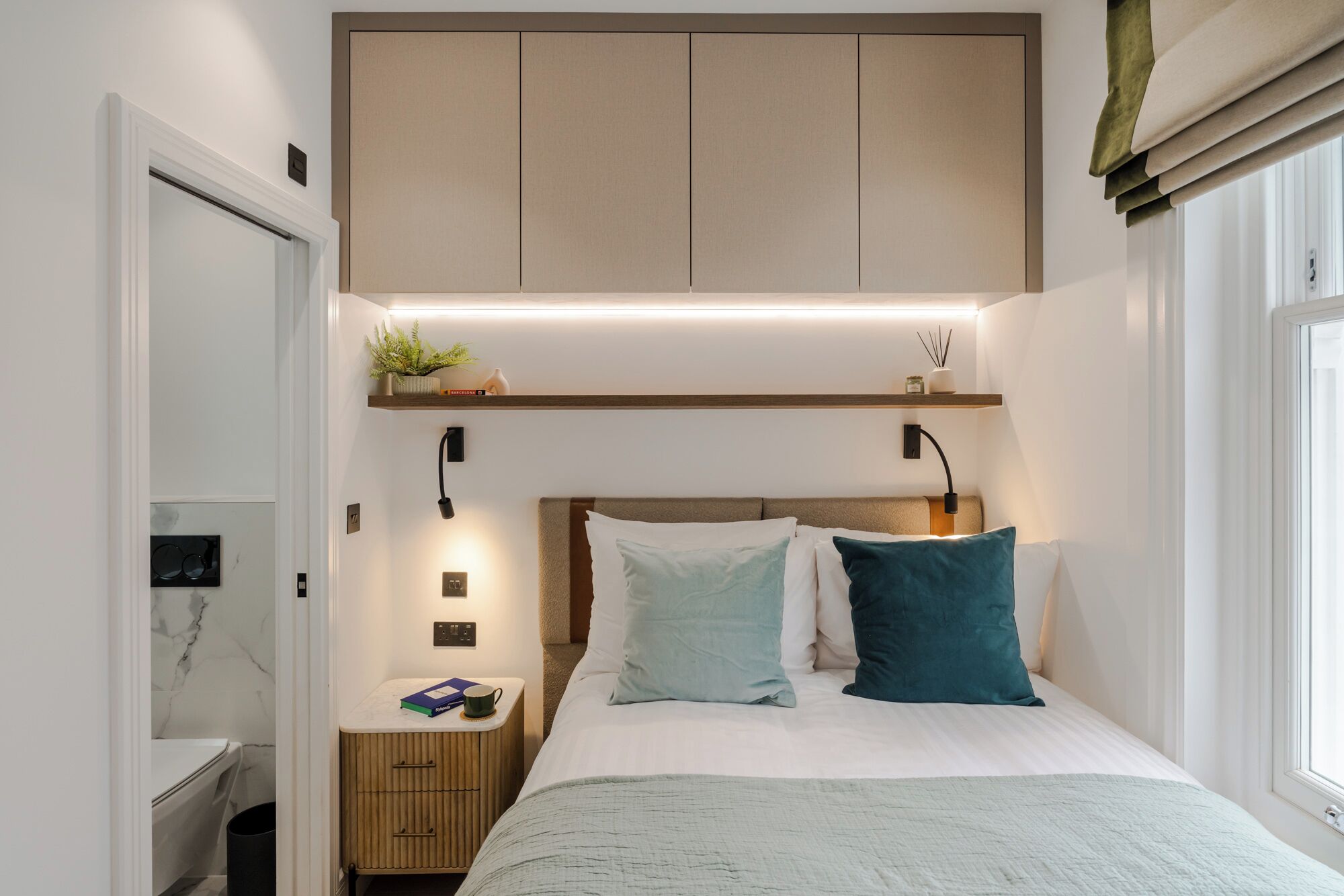 Quarto com cama, mesas de cabeceira, prateleira na parede e janela com cortinas.