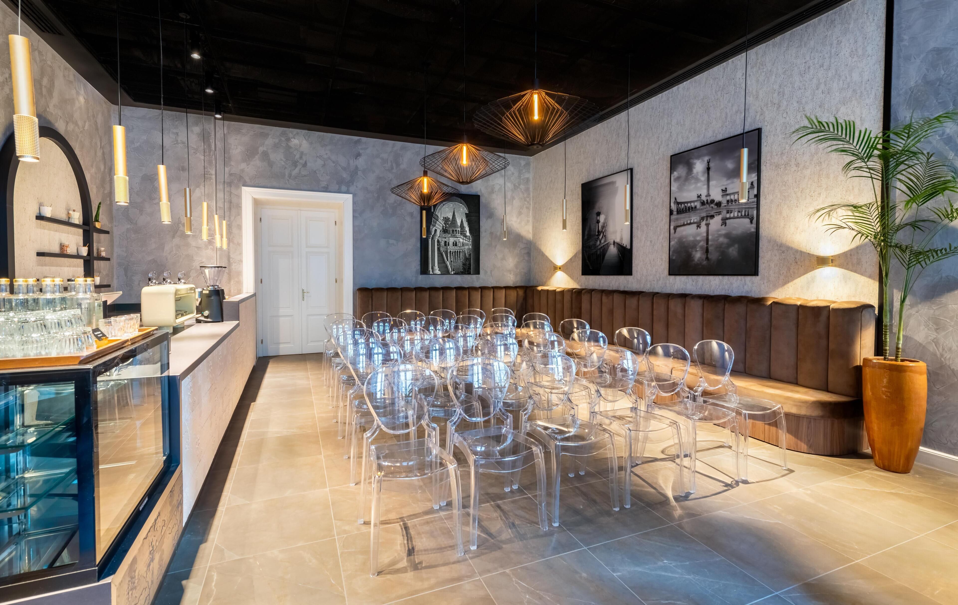 Sala de conferências com cadeiras transparentes dispostas em fileiras, área de buffet com vitrines de vidro e um espaço de estar com um banco.