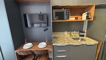 Uma cozinha compacta com micro-ondas, torradeira e pia. Possui uma pequena área de jantar com duas cadeiras e uma mesa.