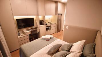 Quarto de hotel com uma cama, escrivaninha com computador, televisão e cozinha compacta.