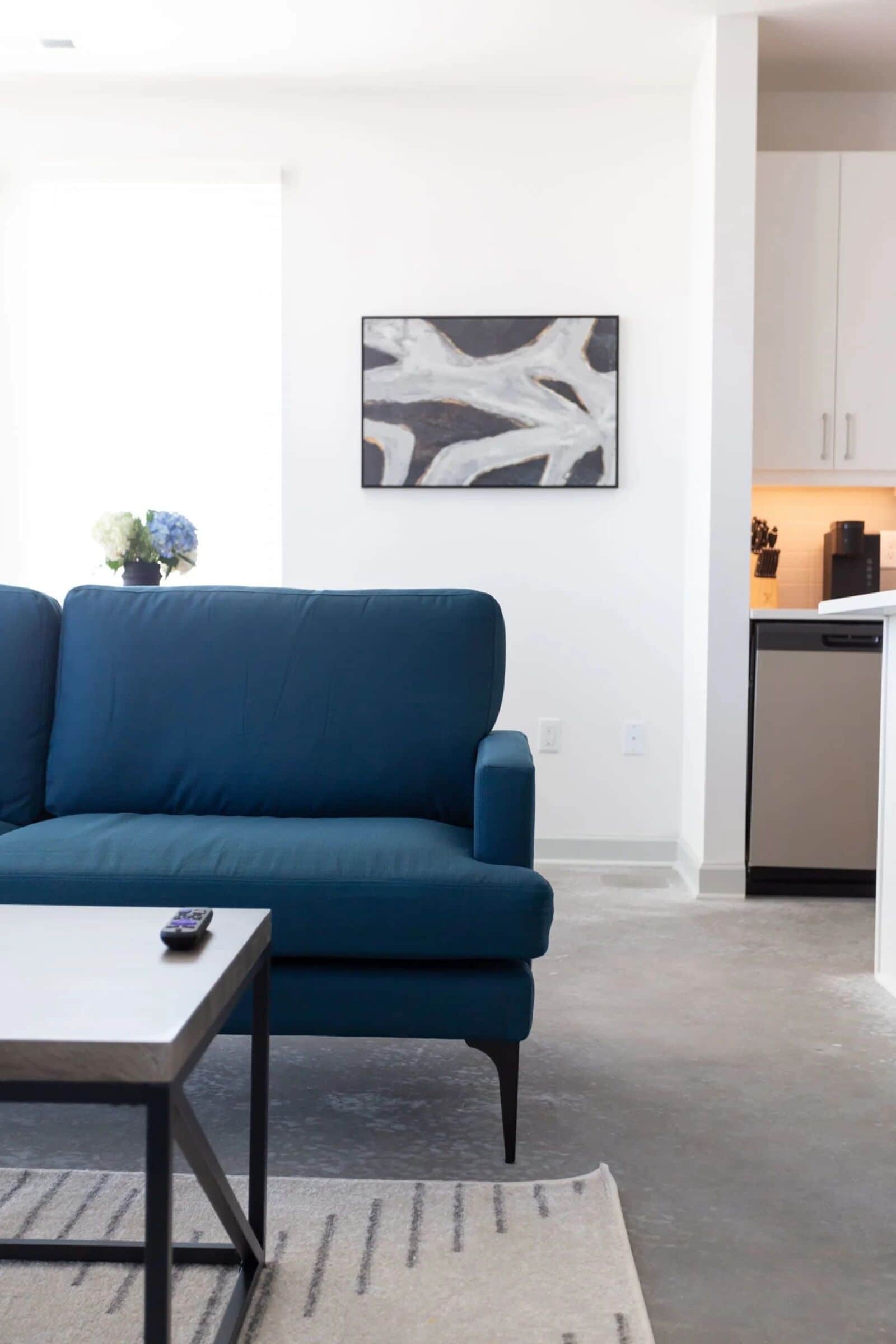 Uma sala de estar moderna com um sofá azul, uma mesinha e um controle remoto sobre ela. Uma cozinha é visível ao fundo.