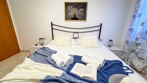 Cama bem arrumada com cobertores brancos e azuis, um livro e um abajur decorativo num criado-mudo.