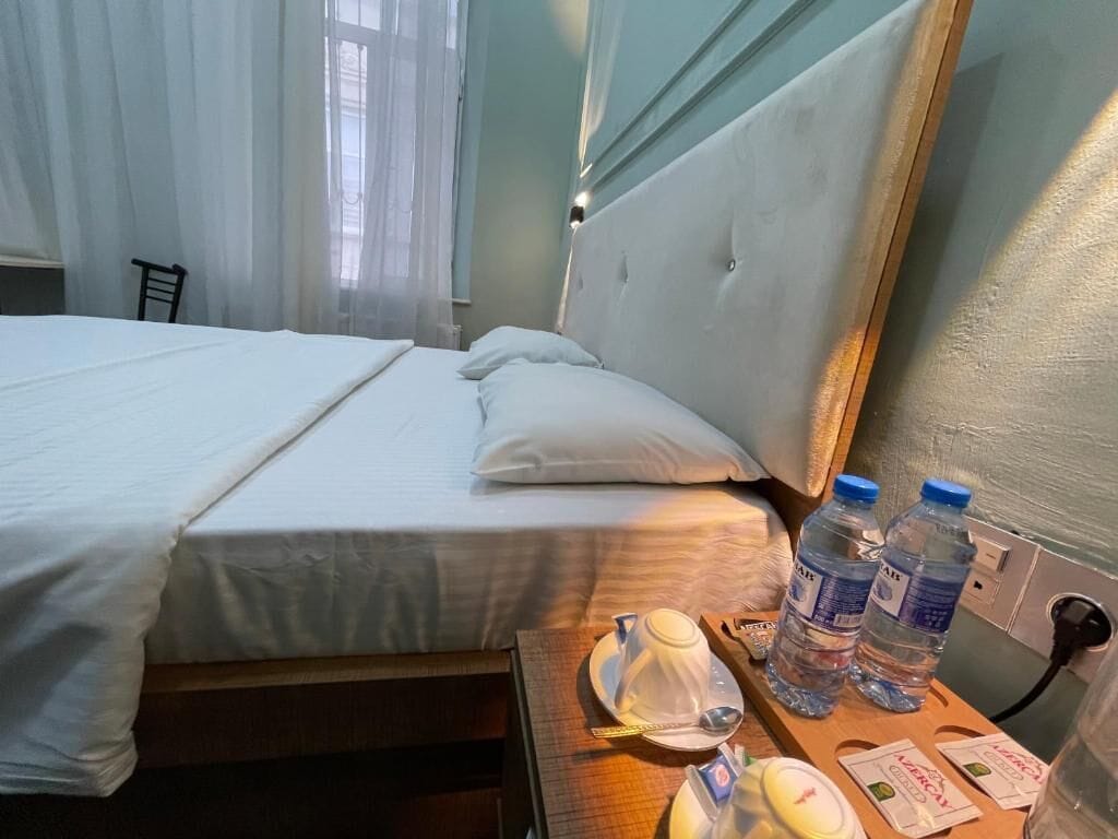 Quarto de hotel com uma cama, uma mesinha de cabeceira com chaleira, garrafas de água e uma bandeja com colher.