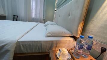 Quarto de hotel com uma cama, uma mesinha de cabeceira com chaleira, garrafas de água e uma bandeja com colher.