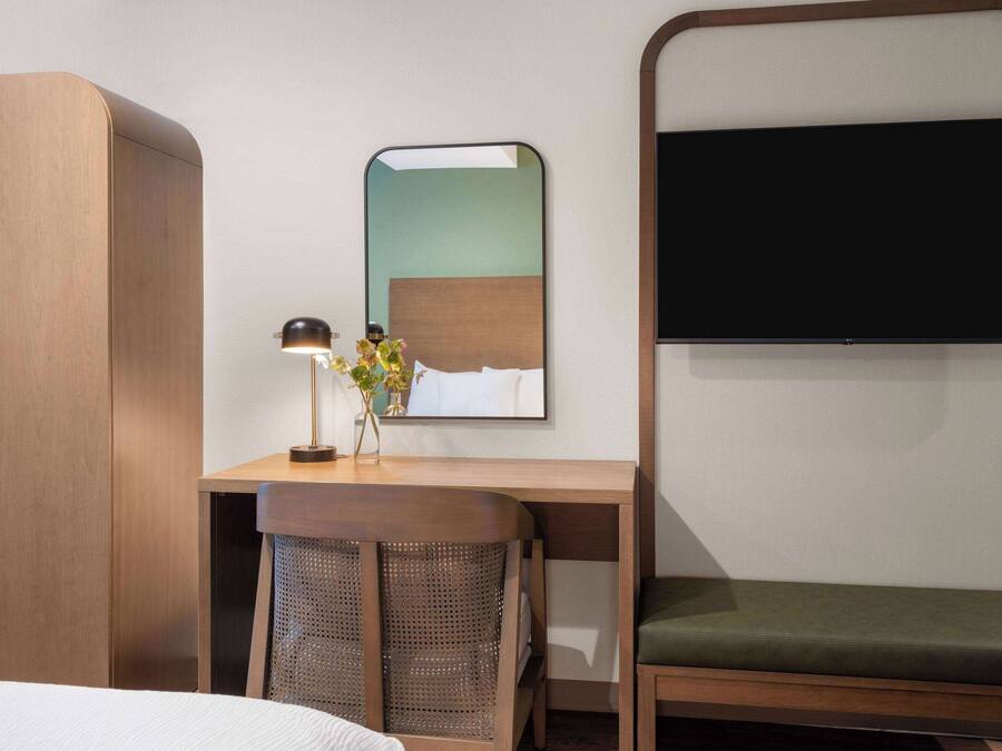 Quarto de hotel com guarda-roupa de madeira, uma escrivaninha com luminária, um espelho, televisão e um banquinho estofado verde.