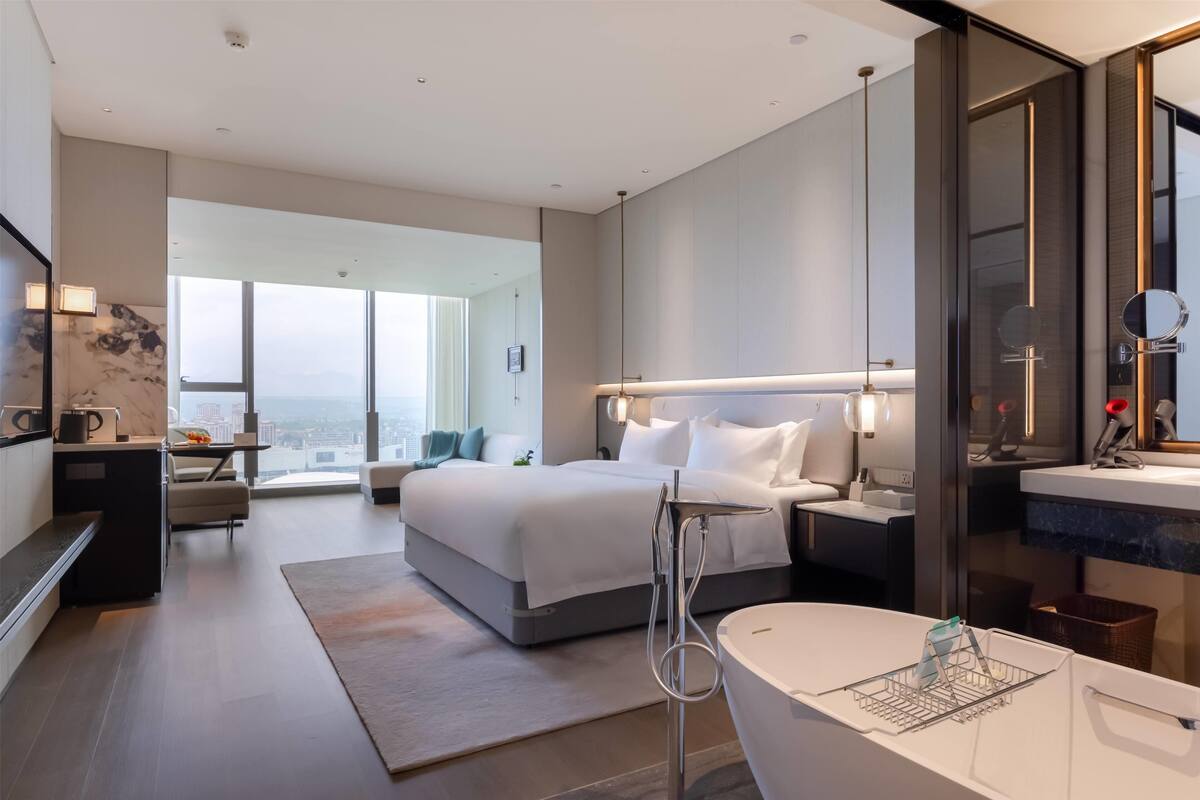 Quarto de hotel moderno com uma cama grande, banheiro com banheira e pia, escrivaninha com cadeira e vista da cidade através de amplas janelas.