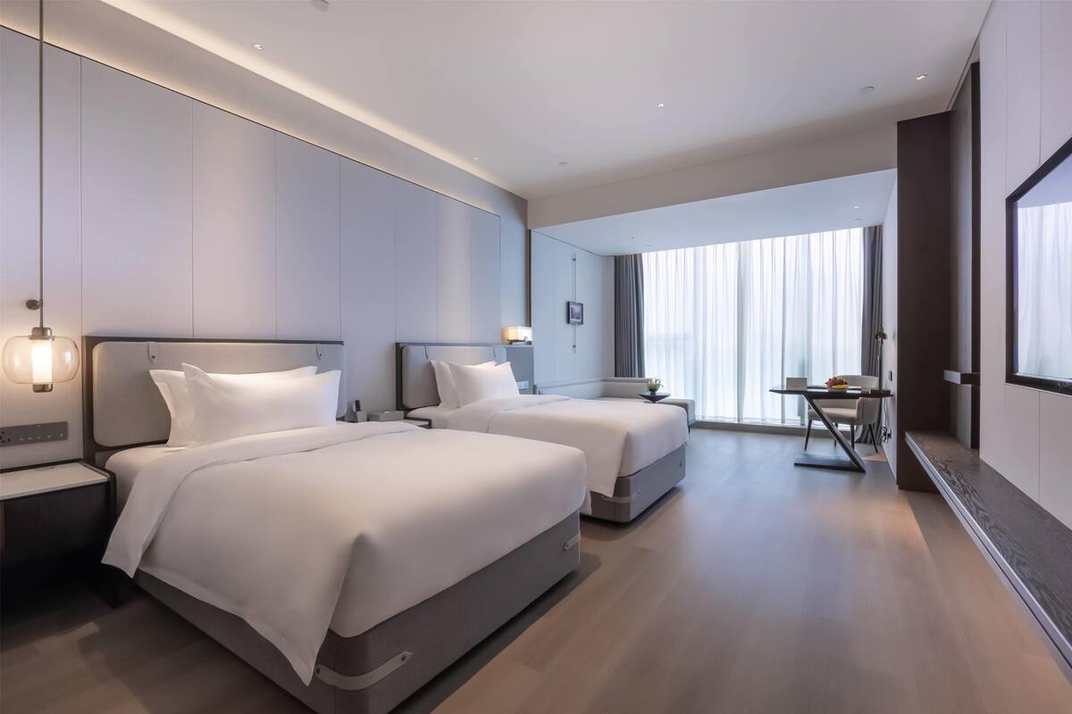 Quarto de hotel moderno com duas camas, uma mesinha e uma janela grande com cortinas.