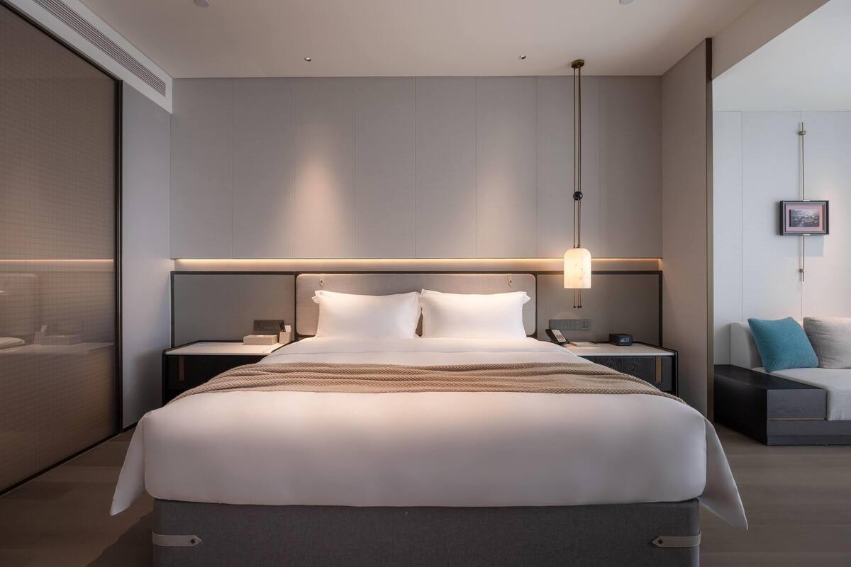 Quarto de hotel moderno com cama grande, mesas de cabeceira, um criado-mudo, luminária na parede e um sofá.