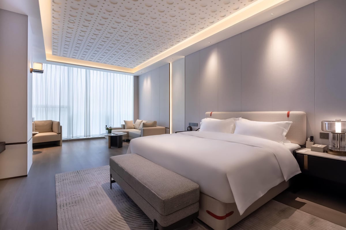 Quarto de hotel moderno com uma cama grande, área de estar com sofá e poltronas, uma mesinha e uma janela ampla com cortinas leves.