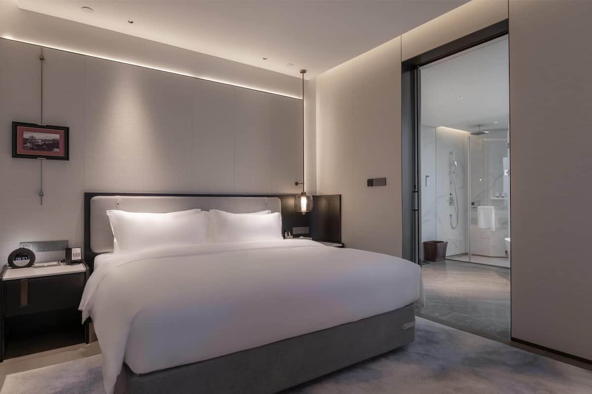 Quarto de hotel moderno com uma cama grande, mesas de cabeceira, um criado-mudo, uma luminária de parede e um quadro emoldurado.