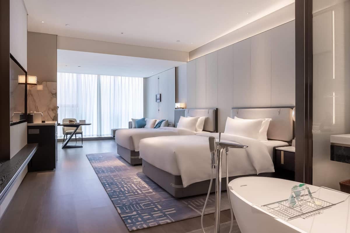 Quarto de hotel moderno com duas camas, área de estar, uma janela grande e banheiro com banheira.