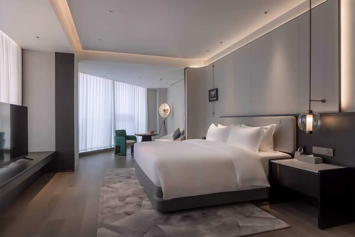 Quarto de hotel moderno com uma cama grande, mesas de cabeceira, televisão e uma janela ampla com persianas.