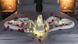 Cama bem arrumada com arranjo de toalha em formato de coração e pétalas de rosa, iluminada por luzes na parede.