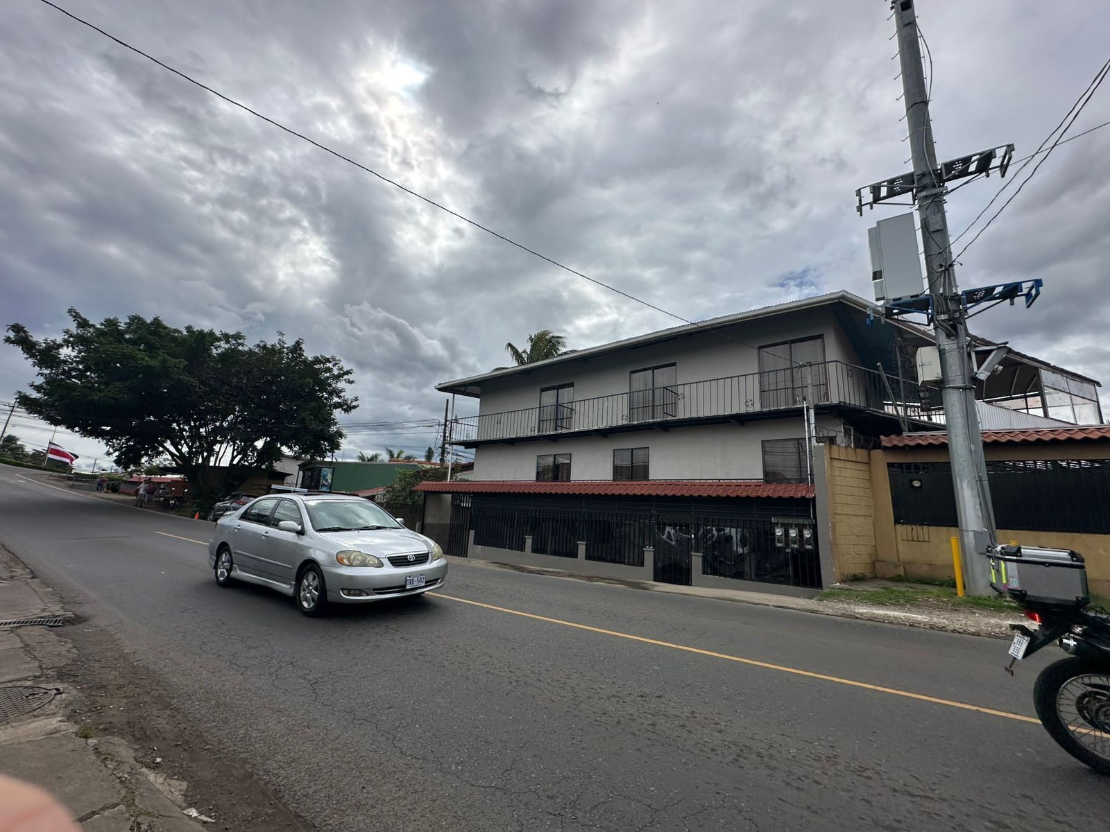 Vista da rua com um prédio de dois andares, um carro estacionado e uma motocicleta.