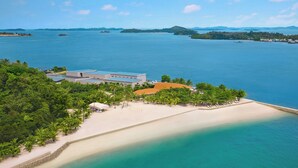 Vista aérea de uma propriedade costeira com praia, um edifício com telhado azul e vegetação ao redor.