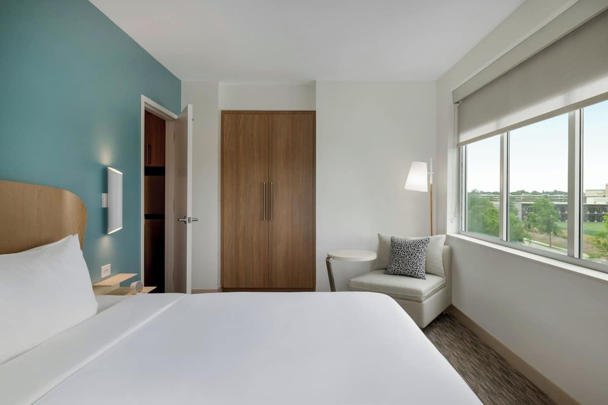 Quarto de hotel com cama, área de estar com luminária, armário e janela com vista.