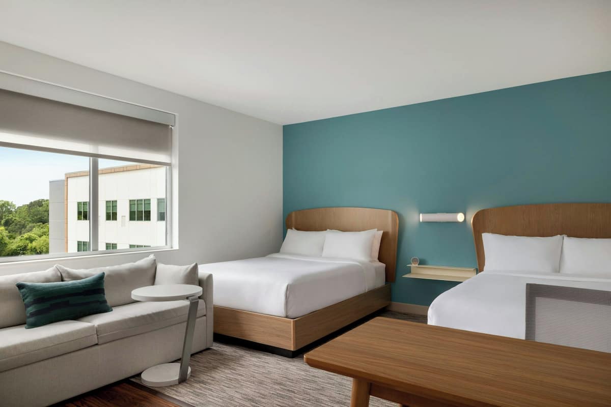 Quarto de hotel moderno com uma grande janela, um sofá, uma cama com cabeceira de madeira e uma pequena mesa de cabeceira.