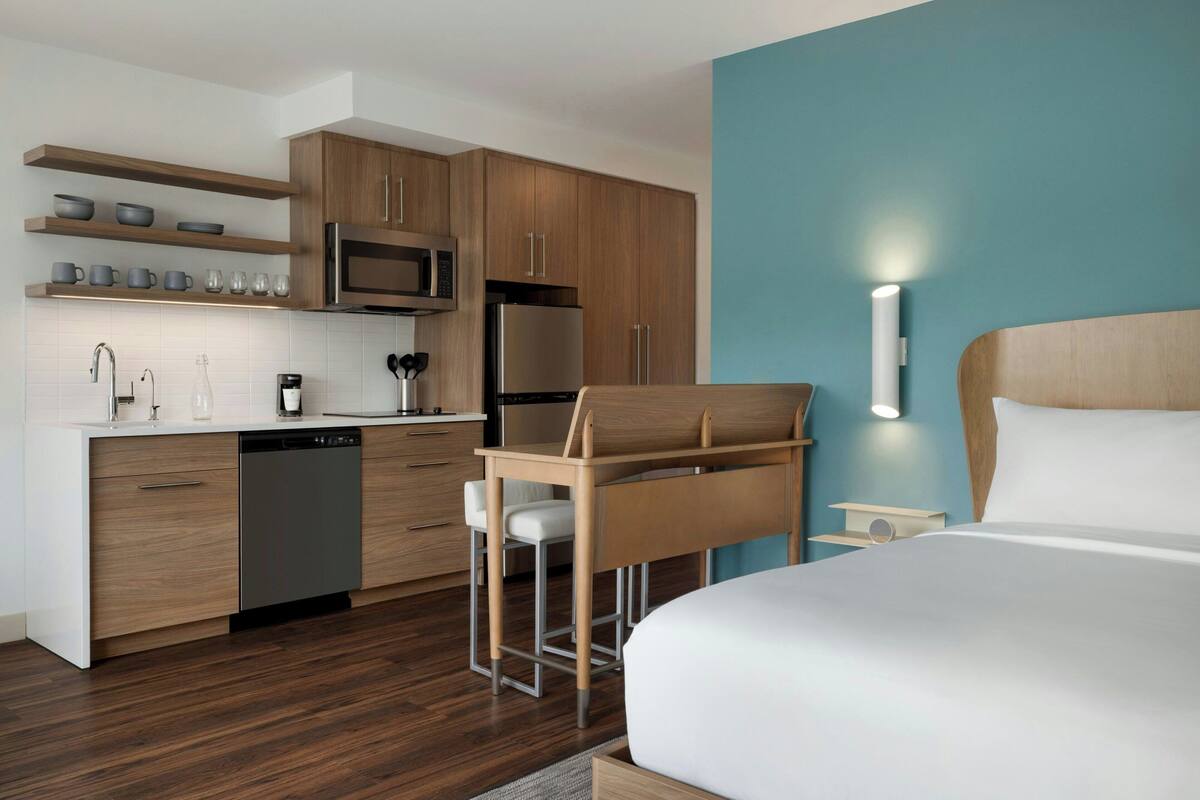 Quarto de hotel moderno com escrivaninha de madeira, uma cama, micro-ondas e frigorífico.