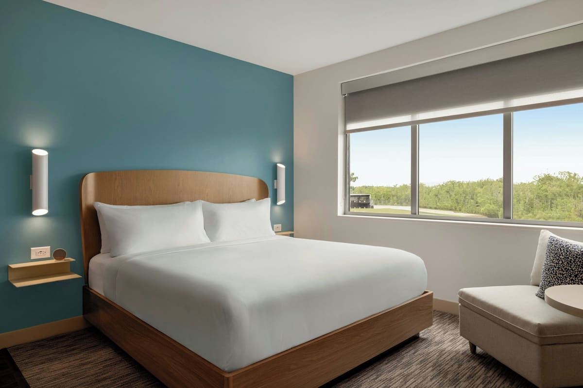 Quarto de hotel moderno com uma cama grande, uma mesinha de cabeceira, uma cadeira e uma janela com vista para vegetação.