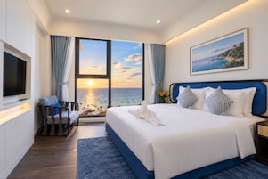 Quarto de hotel com uma cama grande, TV, cadeira e vista para o mar ao pôr do sol.