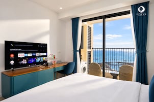 Quarto de hotel com cama, escrivaninha com computador, TV exibindo serviços de streaming, varanda com vista para o mar e janela com cortinas.