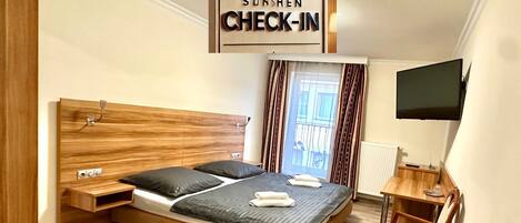 Quarto de hotel com cama, escrivaninha, televisão e janela com cortinas.