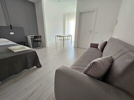 Quarto moderno e bem iluminado, com uma cama, sofá cinza e área de refeições com cadeiras e mesa.