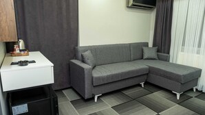 Quarto moderno com sofá seccional cinza, uma pequena mesa branca com um controle remoto, uma chaleira e uma bandeja com lanches.