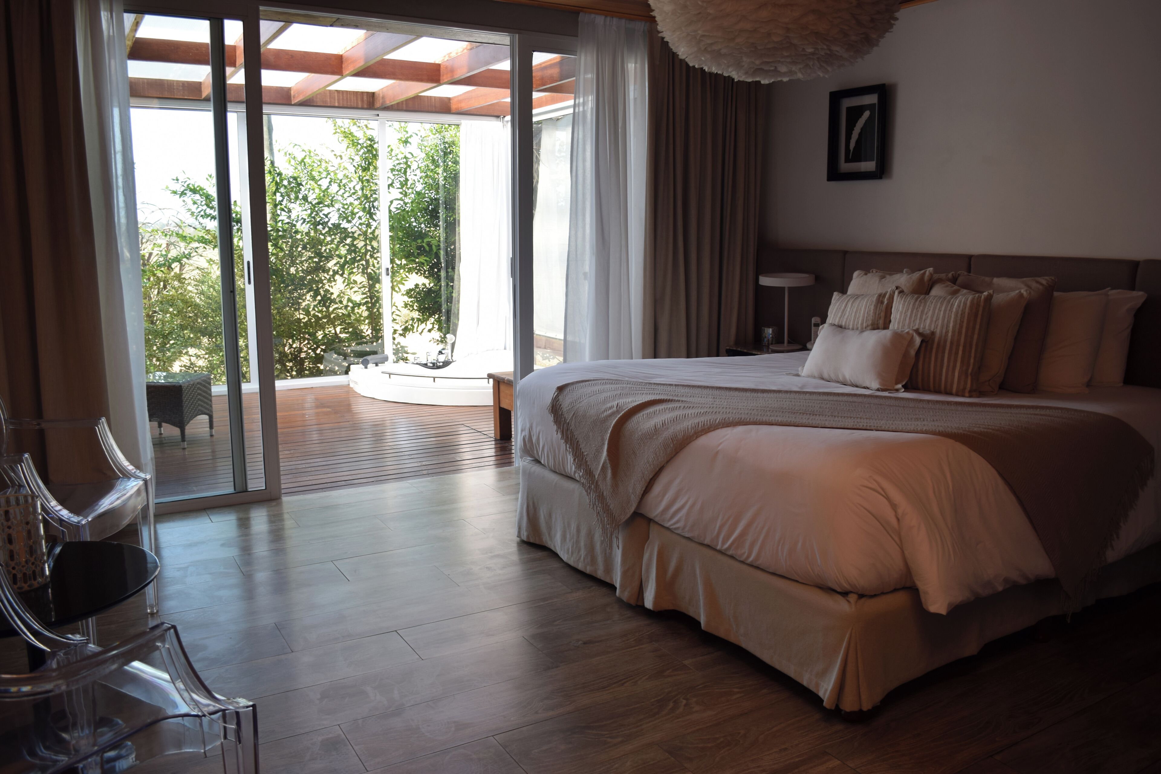 Quarto com uma cama grande, porta de vidro para uma varanda e área de jantar com cadeiras transparentes.