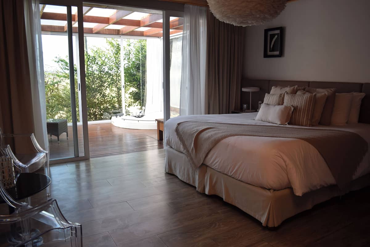 Quarto com uma cama grande, porta de vidro para uma varanda e área de jantar com cadeiras transparentes.