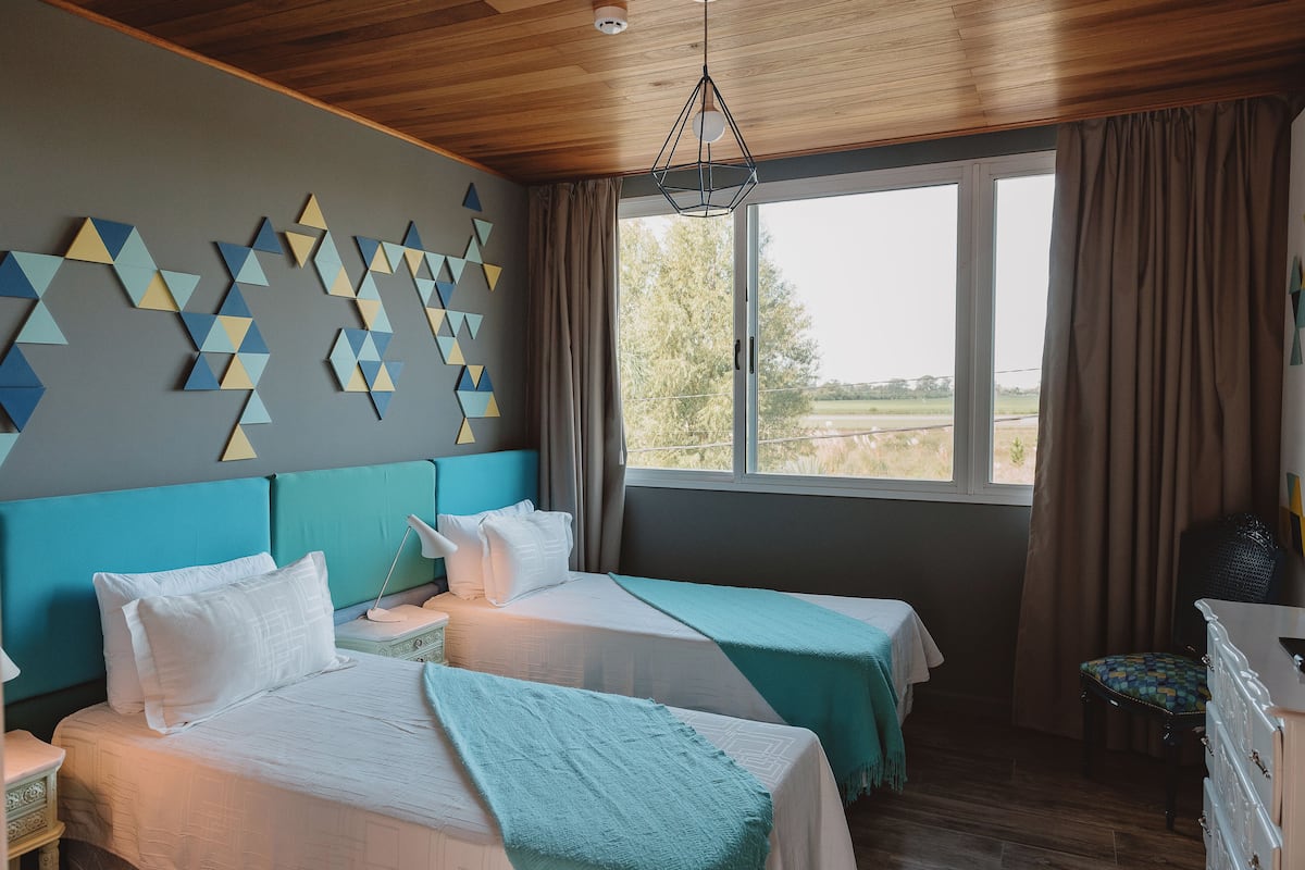 Quarto com duas camas, teto de madeira, janela grande com cortinas e parede com padrões geométricos.