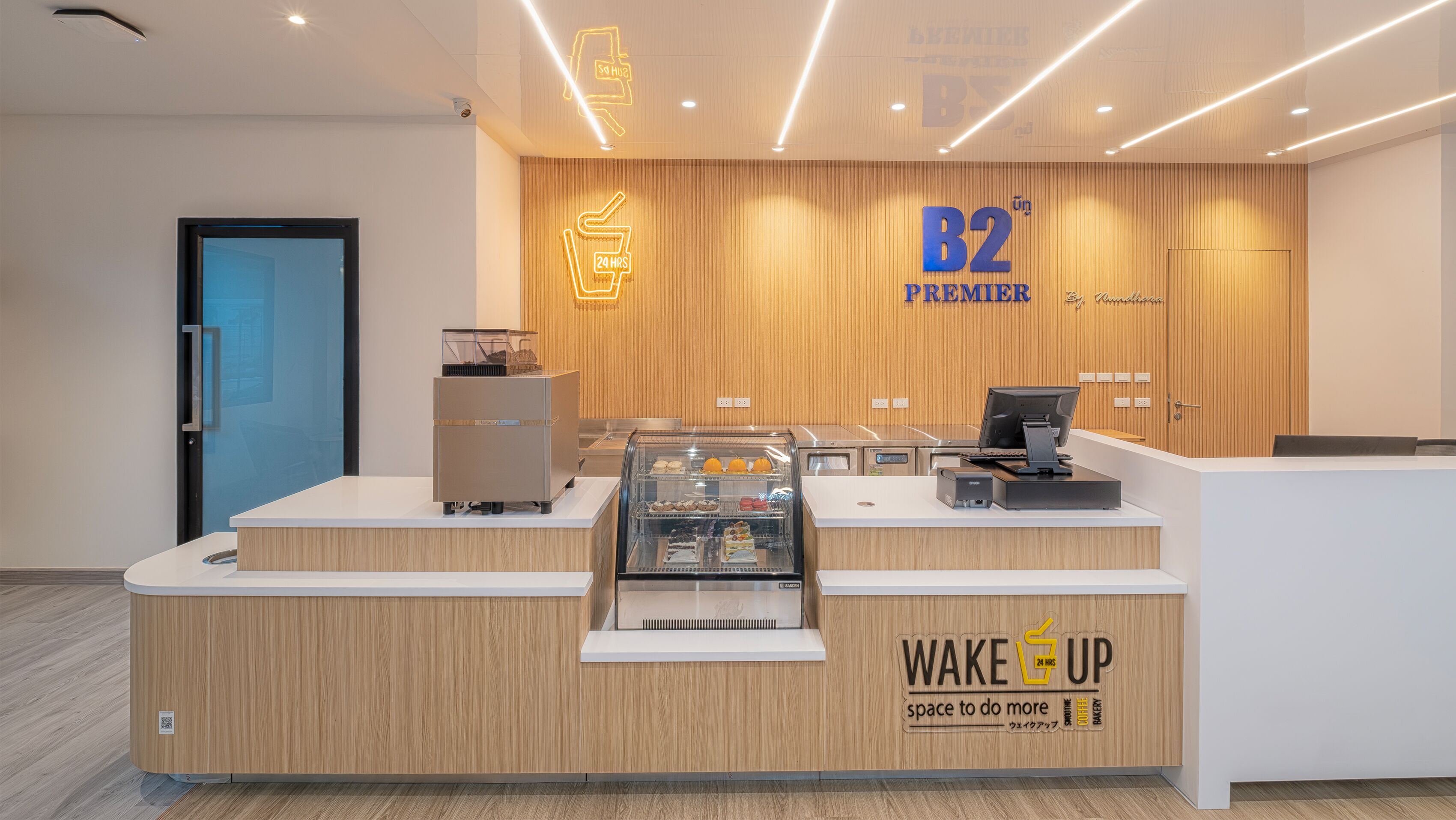 Balcão de recepção de um hotel B2 Premier, com uma máquina de café, uma vitrine de vidro e uma placa com a inscrição 'WAKE UP espaço para fazer mais'.