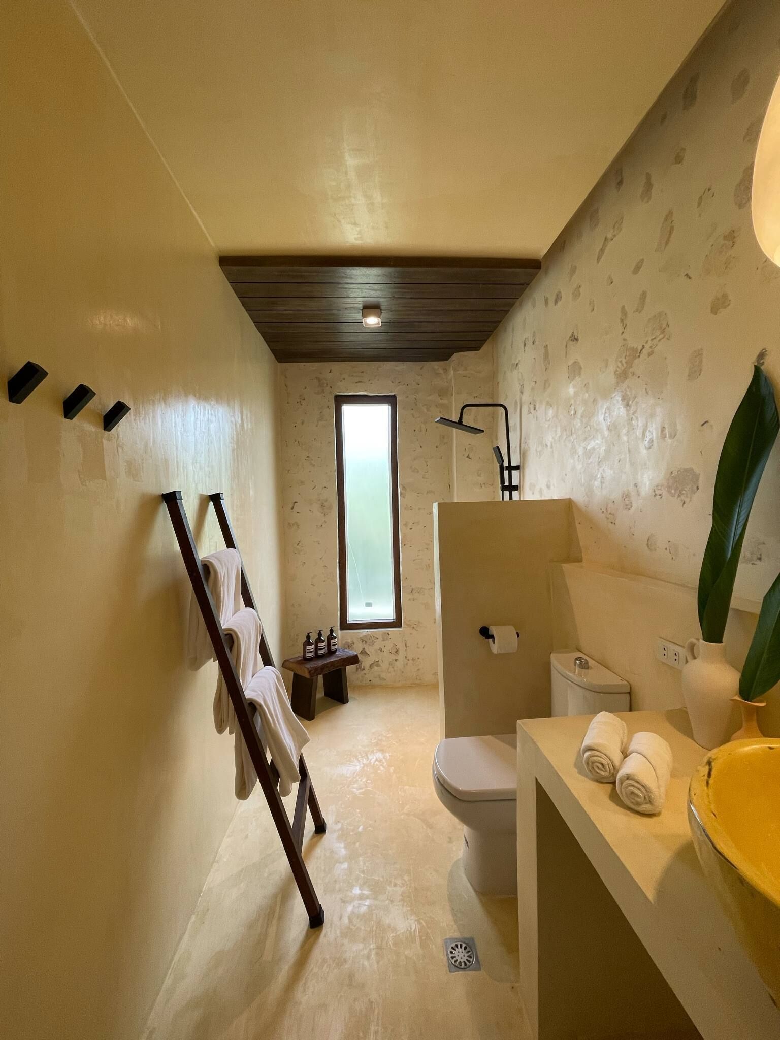 Casa de banho compacta com sanita, lavatório e chuveiro. Há um porta-toalhas e uma pequena mesa com artigos de higiene pessoal. Uma janela permite a entrada de luz natural.
