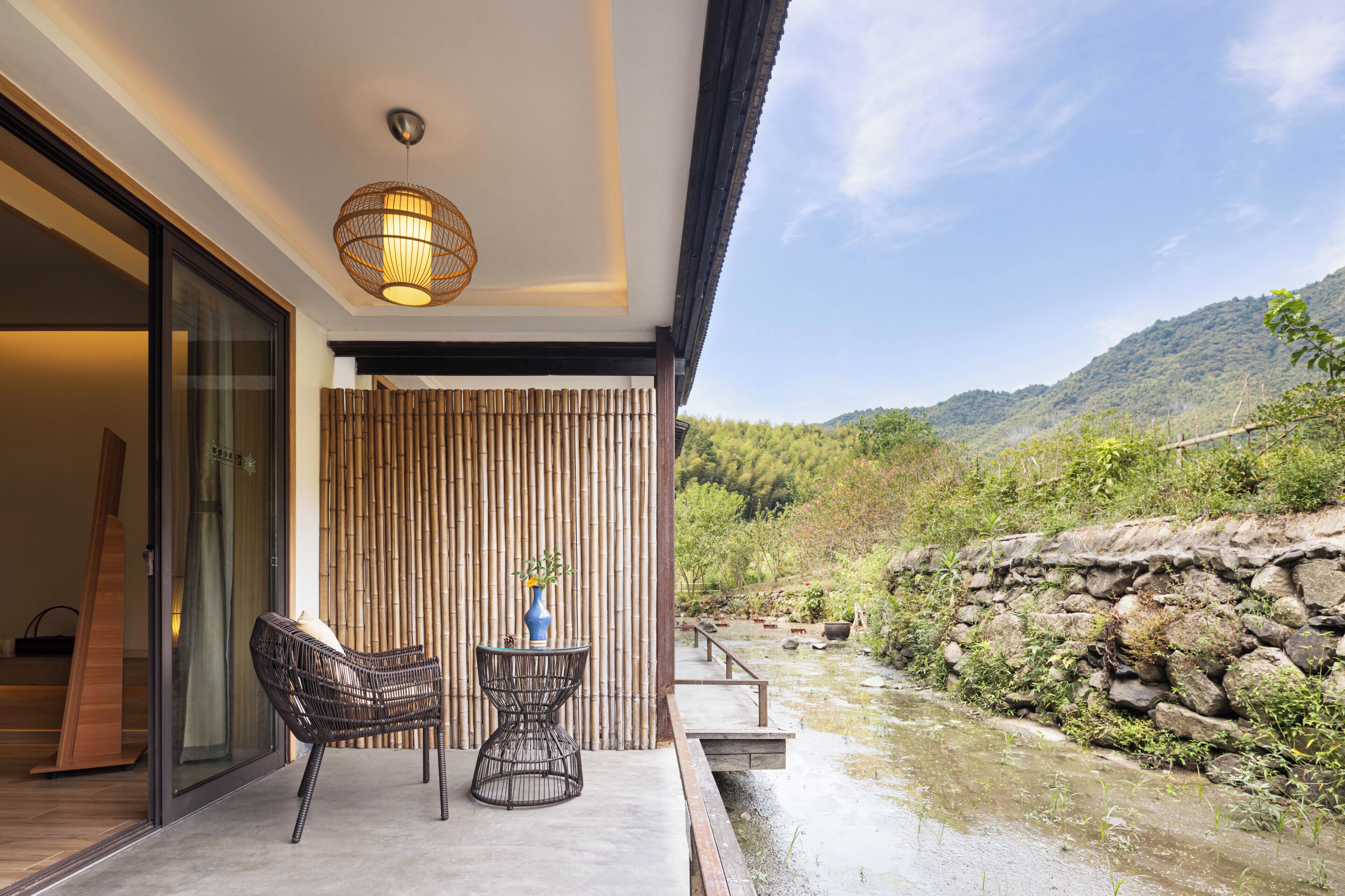 Uma área de estar moderna ao ar livre, com porta de vidro que dá para um espaço interno, parede de bambu e vista de uma paisagem natural.