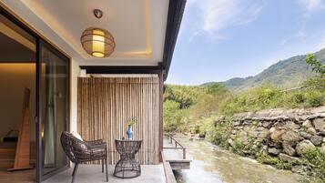 Uma área de estar moderna ao ar livre, com porta de vidro que dá para um espaço interno, parede de bambu e vista de uma paisagem natural.