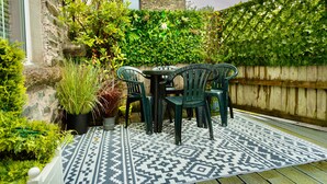 Um pátio com mesa e cadeiras, rodeado por plantas em vasos e um tapete estampado.