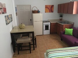 Uma cozinha compacta com geladeira, fogão e pia. Há uma mesa de jantar com um banco e um sofá roxo com travesseiro verde.