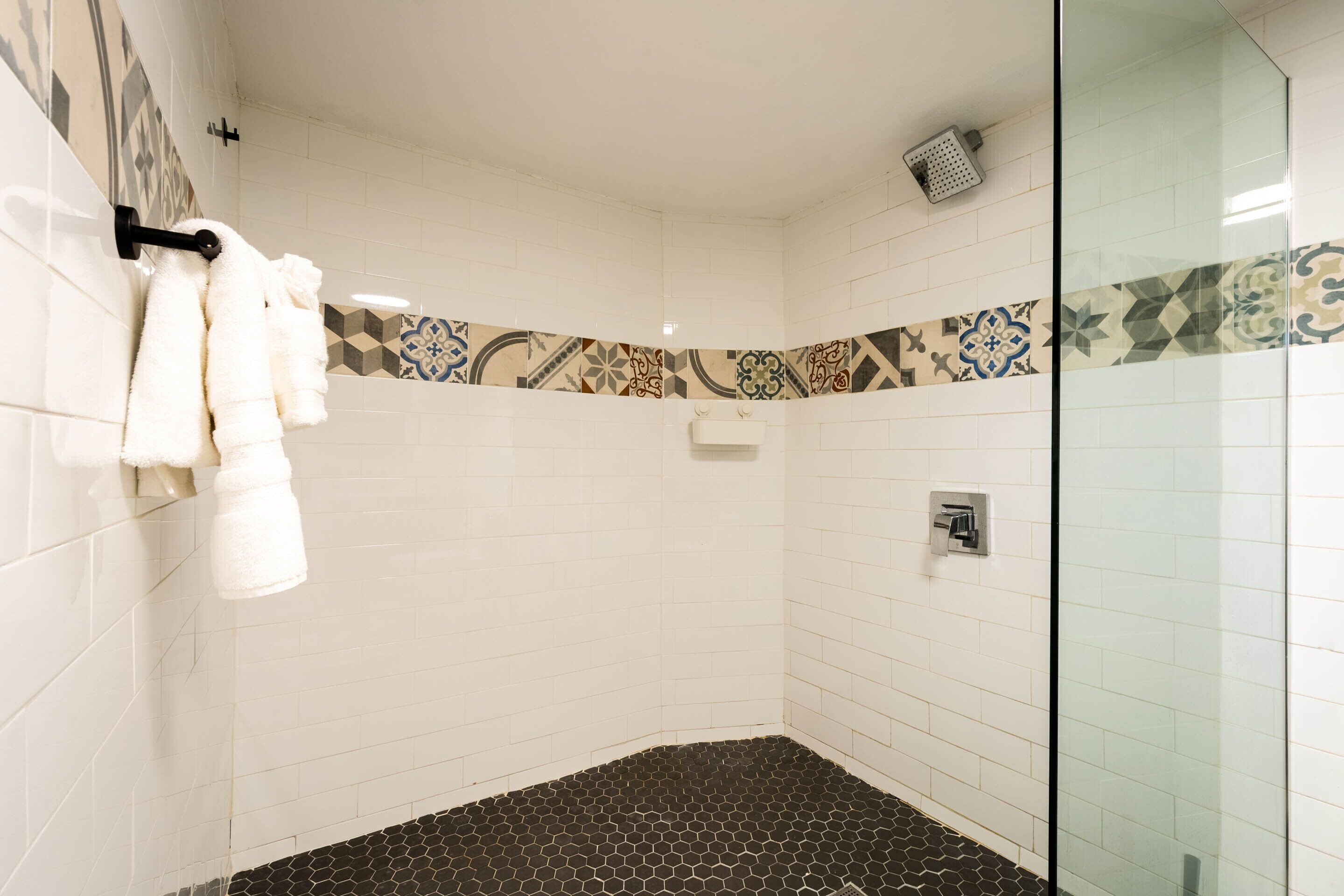 Uma casa de banho de hotel com paredes revestidas de azulejos brancos, cabine de duche em vidro e chão em mosaico hexagonal preto.
