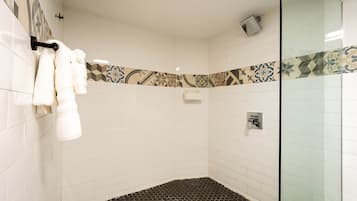 Uma casa de banho de hotel com paredes revestidas de azulejos brancos, cabine de duche em vidro e chão em mosaico hexagonal preto.