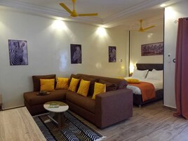 Uma sala de estar moderna com um sofá marrom, uma mesa de centro redonda e uma cama com manta laranja. Há ventiladores de teto, obras de arte nas paredes e um tapete estampado.