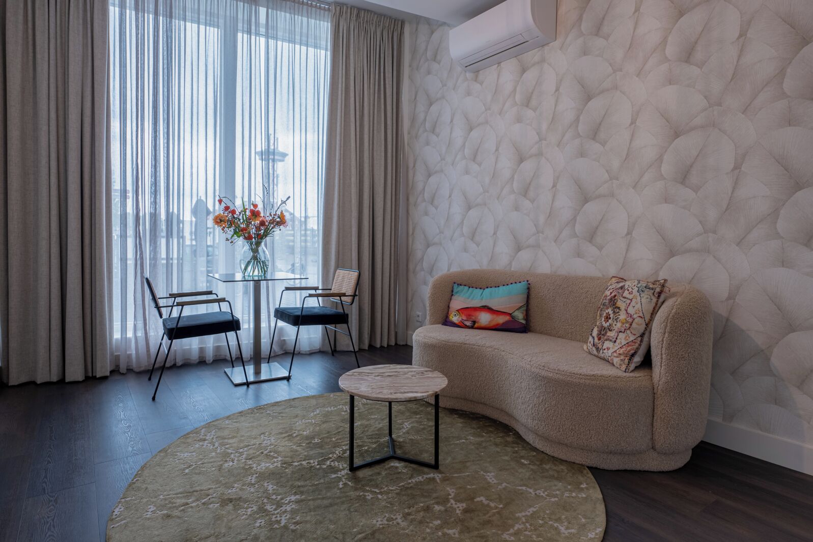 Uma sala de estar moderna com um sofá bege, duas cadeiras pretas, uma mesa de vidro e uma mesa de centro redonda. Um vaso decorativo com flores está sobre a mesa, e o quarto possui uma parede texturizada e uma janela grande com cortinas leves.