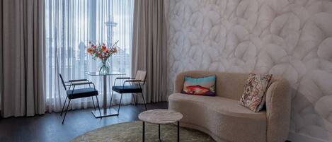 Uma sala de estar moderna com um sofá bege, duas cadeiras pretas, uma mesa de vidro e uma mesa de centro redonda. Um vaso decorativo com flores está sobre a mesa, e o quarto possui uma parede texturizada e uma janela grande com cortinas leves.