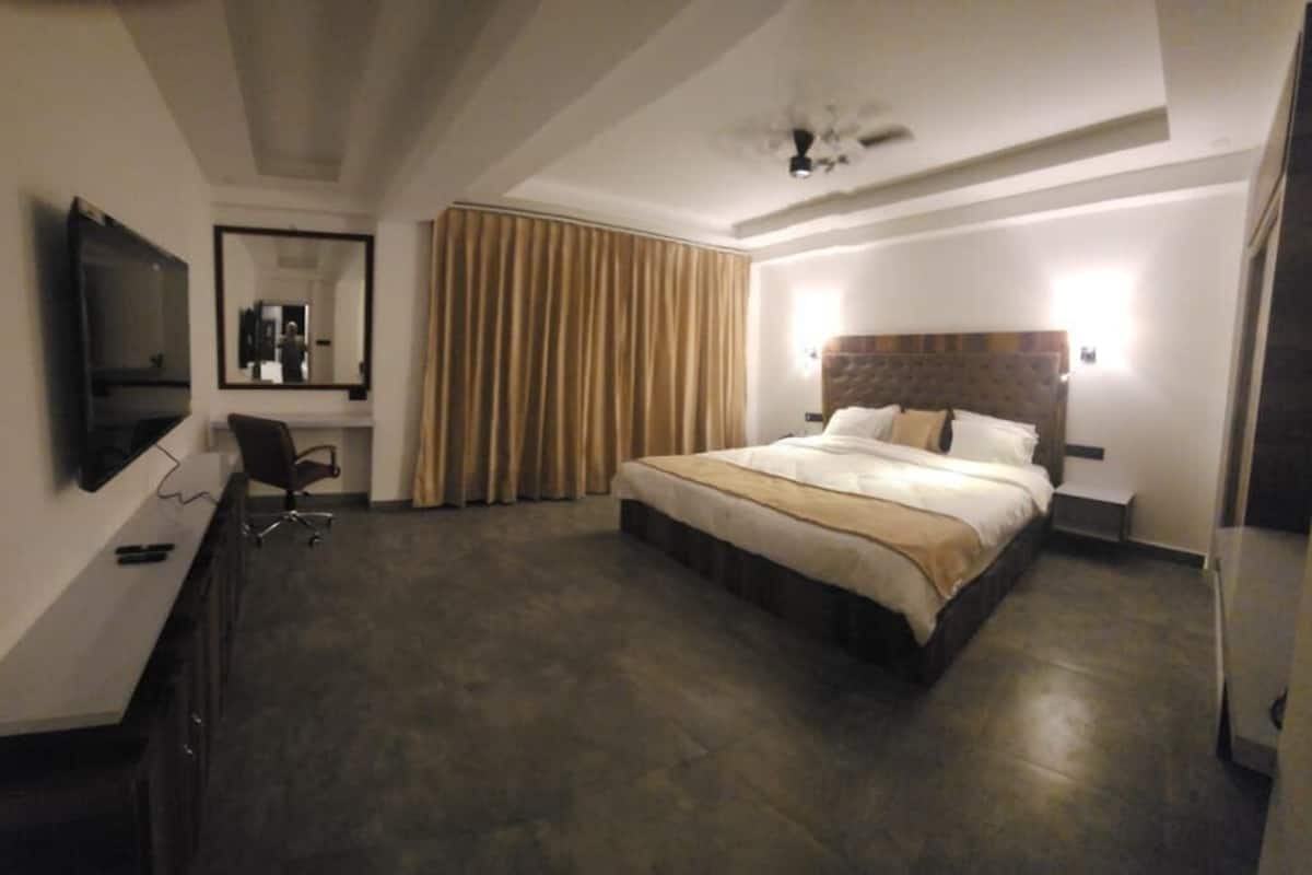 Quarto de hotel com cama, escrivaninha, cadeira, televisão e uma grande janela com cortinas.